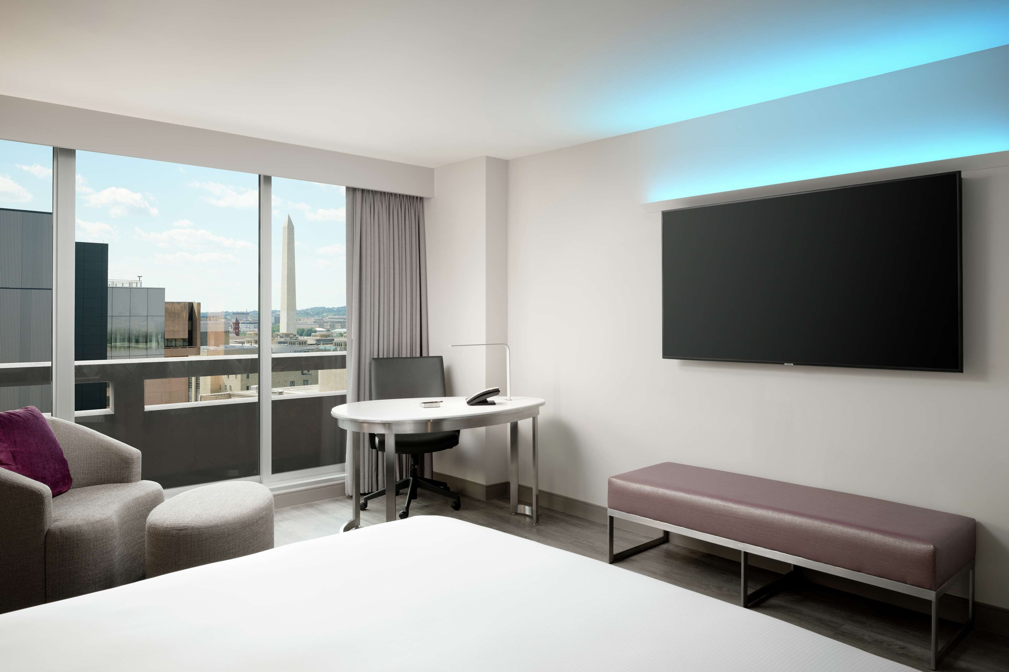 Quarto de hotel moderno com uma cama grande, uma escrivaninha, uma cadeira, um banco, uma TV e vista para a cidade.