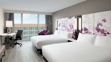 Quarto de hotel com duas camas, uma escrivaninha, uma cadeira e uma janela grande com vista para a cidade.