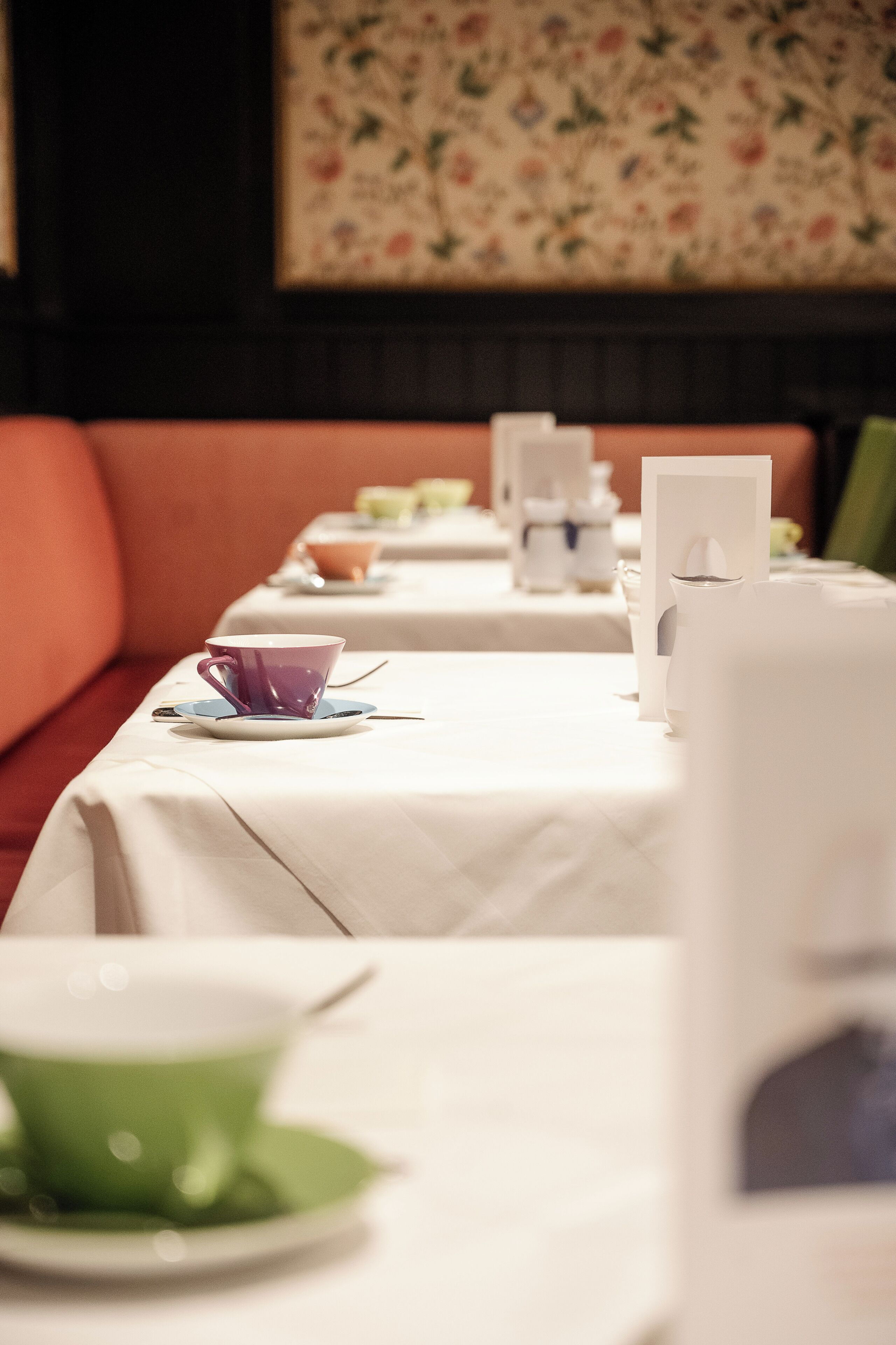 Um ambiente de restaurante com mesas preparadas para refeições, incluindo uma chávena roxa e uma chávena verde em pires.