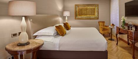 Quarto de hotel com cama, abajures, escrivaninha com cadeira, televisão e um objeto decorativo na parede.