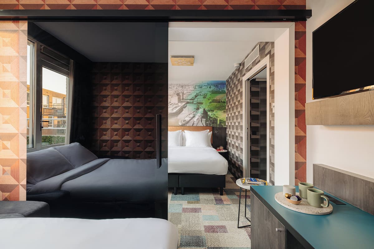 Quarto de hotel com cama, sofá, pequena mesa com bandeja e vista para a cidade através de uma janela.
