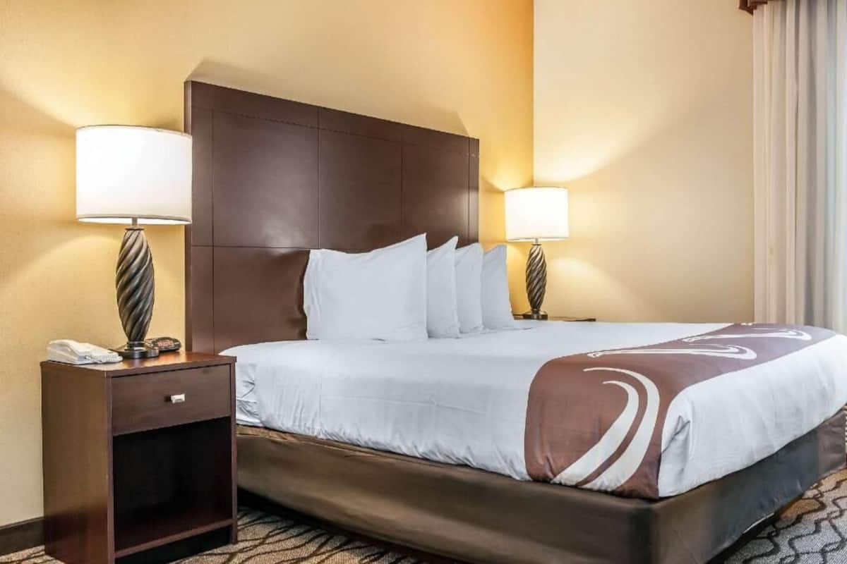 Quarto de hotel com uma cama grande, duas luminárias de cabeceira, uma mesinha de cabeceira com telefone e um tapete estampado.