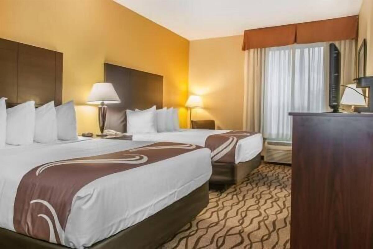 Quarto de hotel com duas camas, uma escrivaninha, uma luminária e uma janela grande com cortinas.