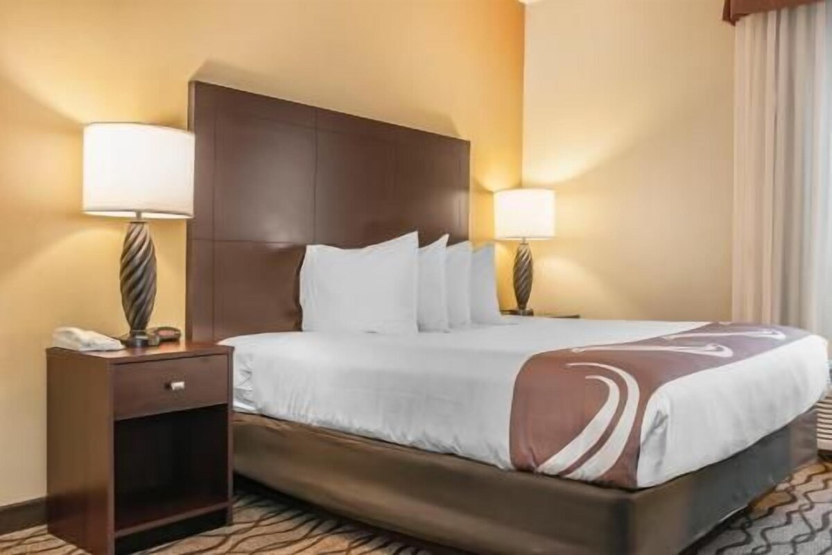 Quarto de hotel com uma cama grande, duas luminárias de cabeceira, uma mesinha de cabeceira e um tapete estampado.