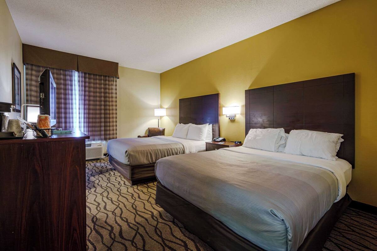 Quarto de hotel com duas camas, uma escrivaninha, um espelho e uma janela com cortinas.