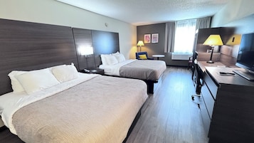 Quarto de hotel com duas camas, uma escrivaninha com televisão, uma luminária e uma pequena mesa.