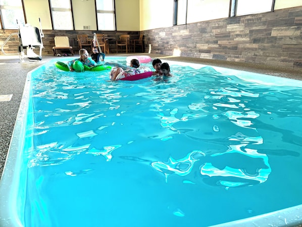Piscina interior com água cristalina, cadeiras de madeira e parede de pedra.