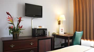 Quarto de hotel com uma cama, uma escrivaninha com telefone, micro-ondas, televisão, luminária e uma planta.