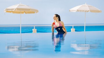 Mulher em piscina com dois guarda-sóis brancos, céu azul claro e mar calmo ao fundo.