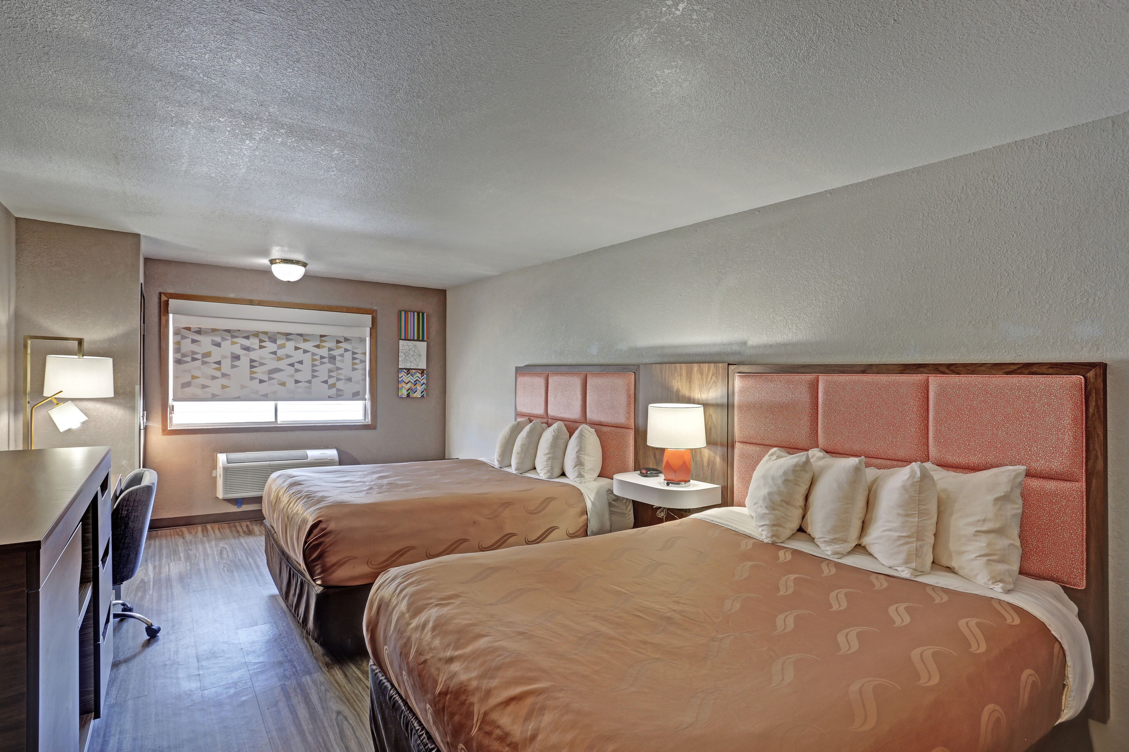 Quarto de hotel com duas camas, uma escrivaninha, uma cadeira e uma janela com cortinas geométricas.