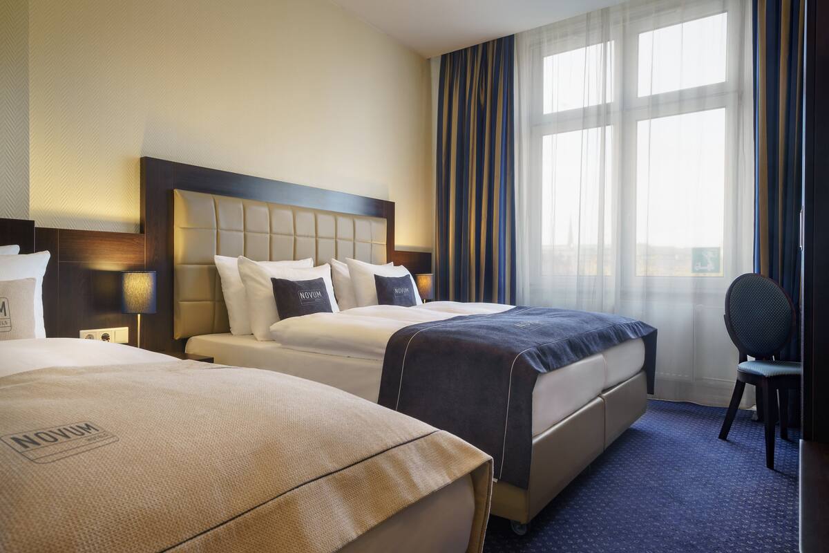 Quarto de hotel com duas camas, uma cadeira e uma janela grande com cortinas.