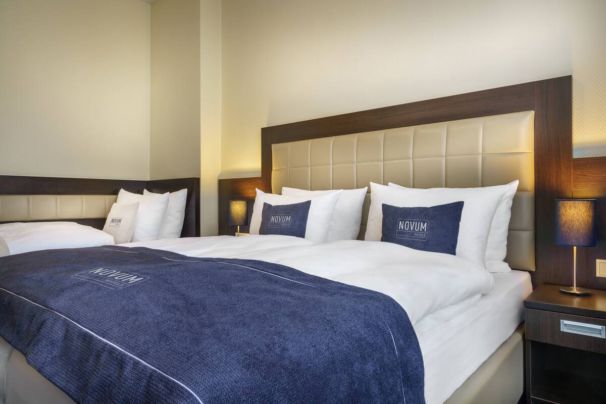 Quarto de hotel com uma cama grande, várias almofadas, uma mesa de cabeceira com abajur e um cobertor azul-escuro.
