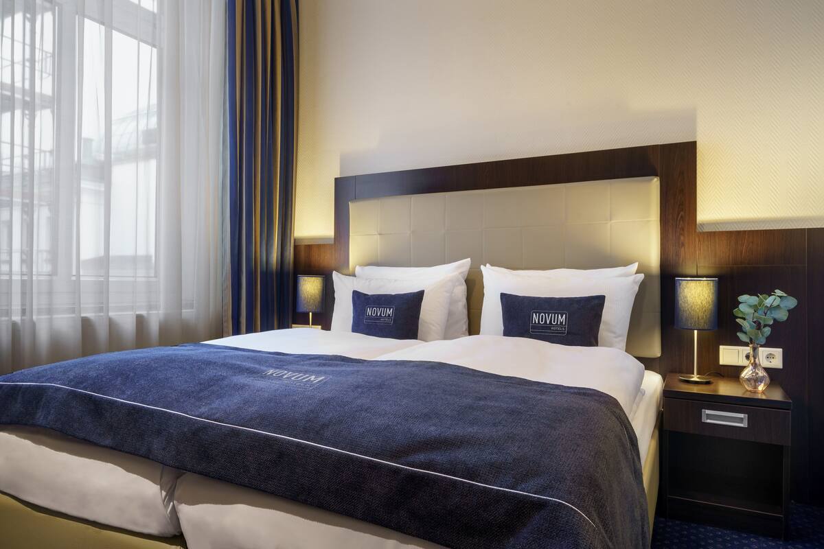 Quarto de hotel com uma cama grande, duas mesas de cabeceira e uma janela com cortinas finas.