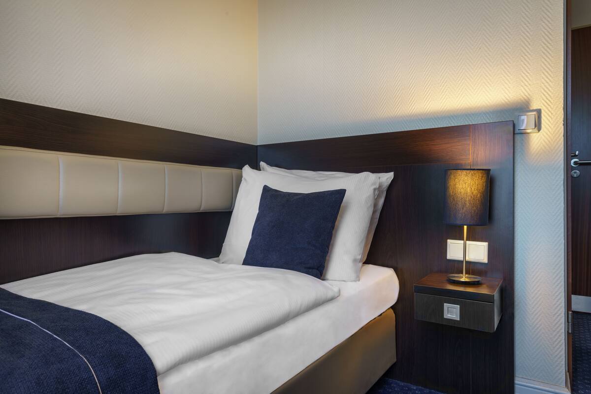 Quarto de hotel com cama, mesa de cabeceira com abajur e interruptor de luz na parede.