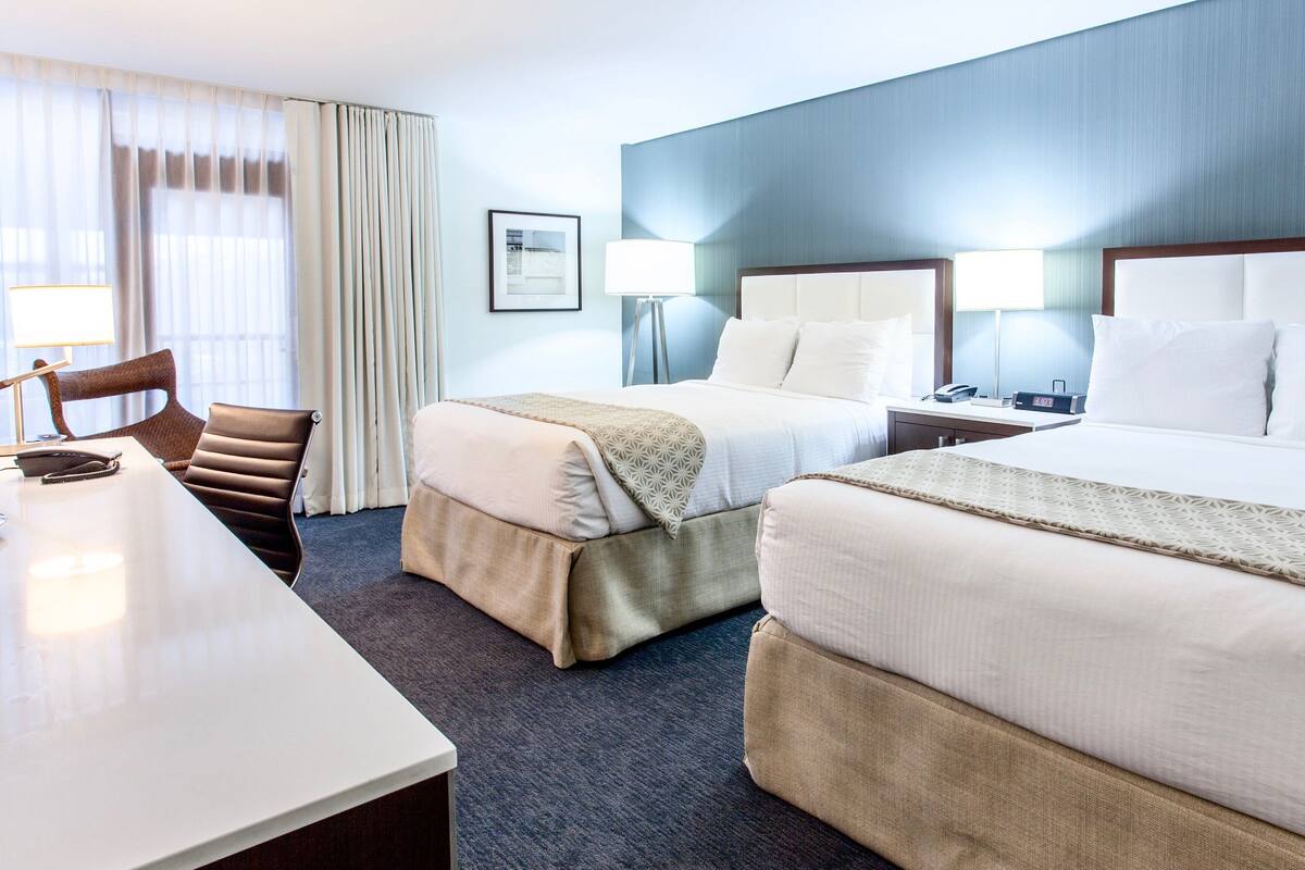 Quarto de hotel com duas camas, uma escrivaninha, uma cadeira e um abajur.