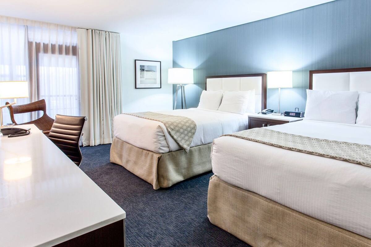 Quarto de hotel com duas camas, uma mesa, uma cadeira, um abajur e um quadro na parede.