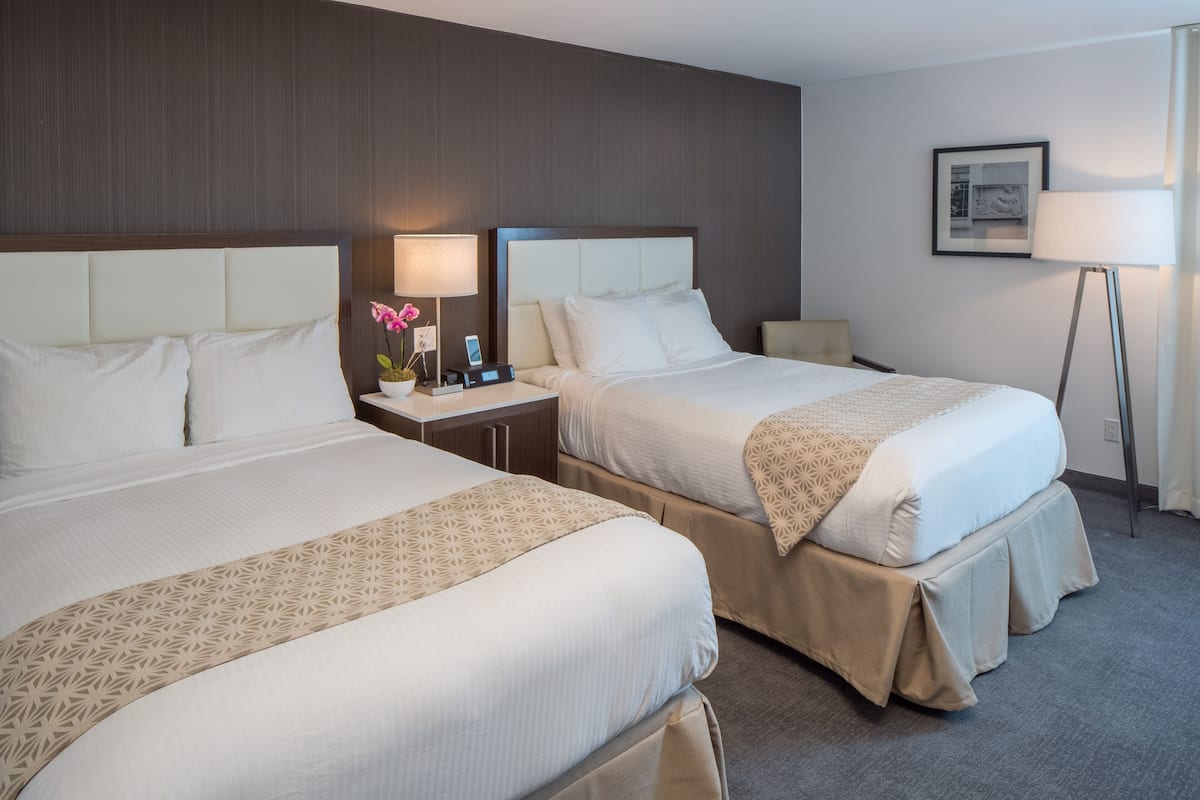 Quarto de hotel com duas camas, uma mesinha de cabeceira com abajur, um quadro na parede e um candeeiro de chão.