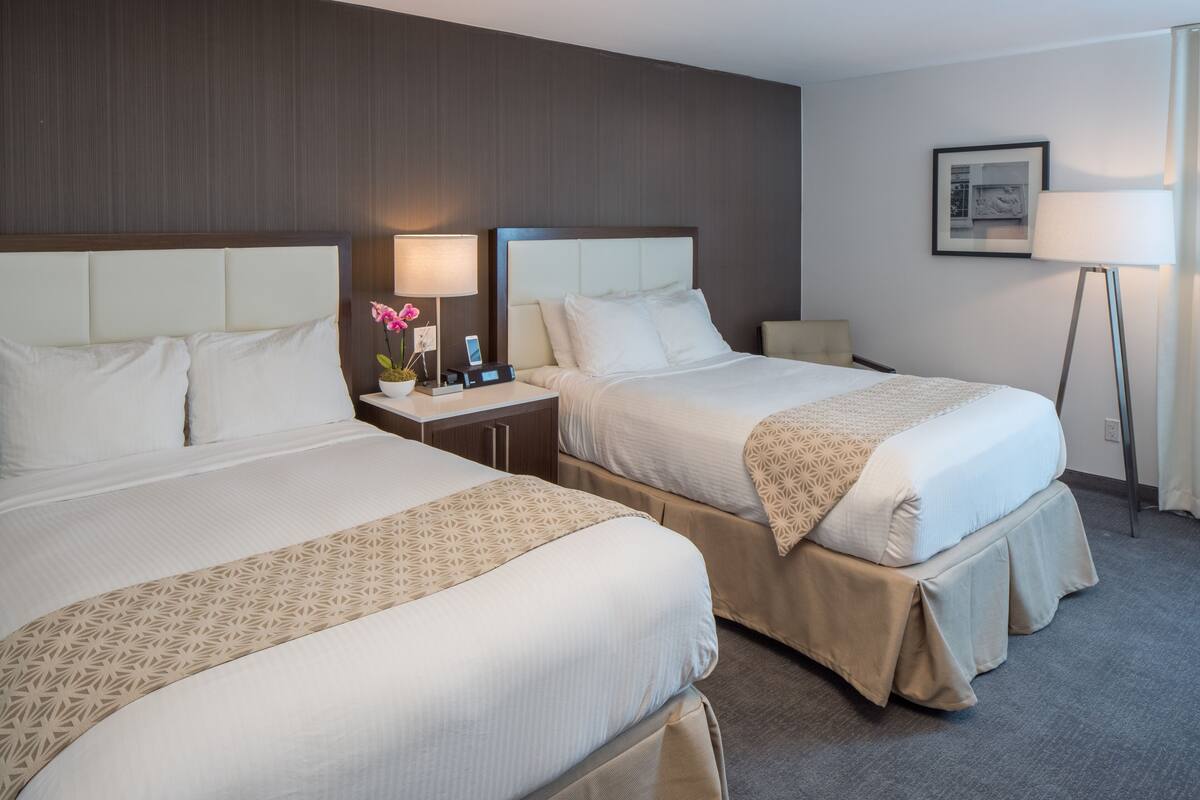 Quarto de hotel com duas camas, uma mesinha de cabeceira com abajur, um quadro pendurado na parede e um candeeiro de chão.