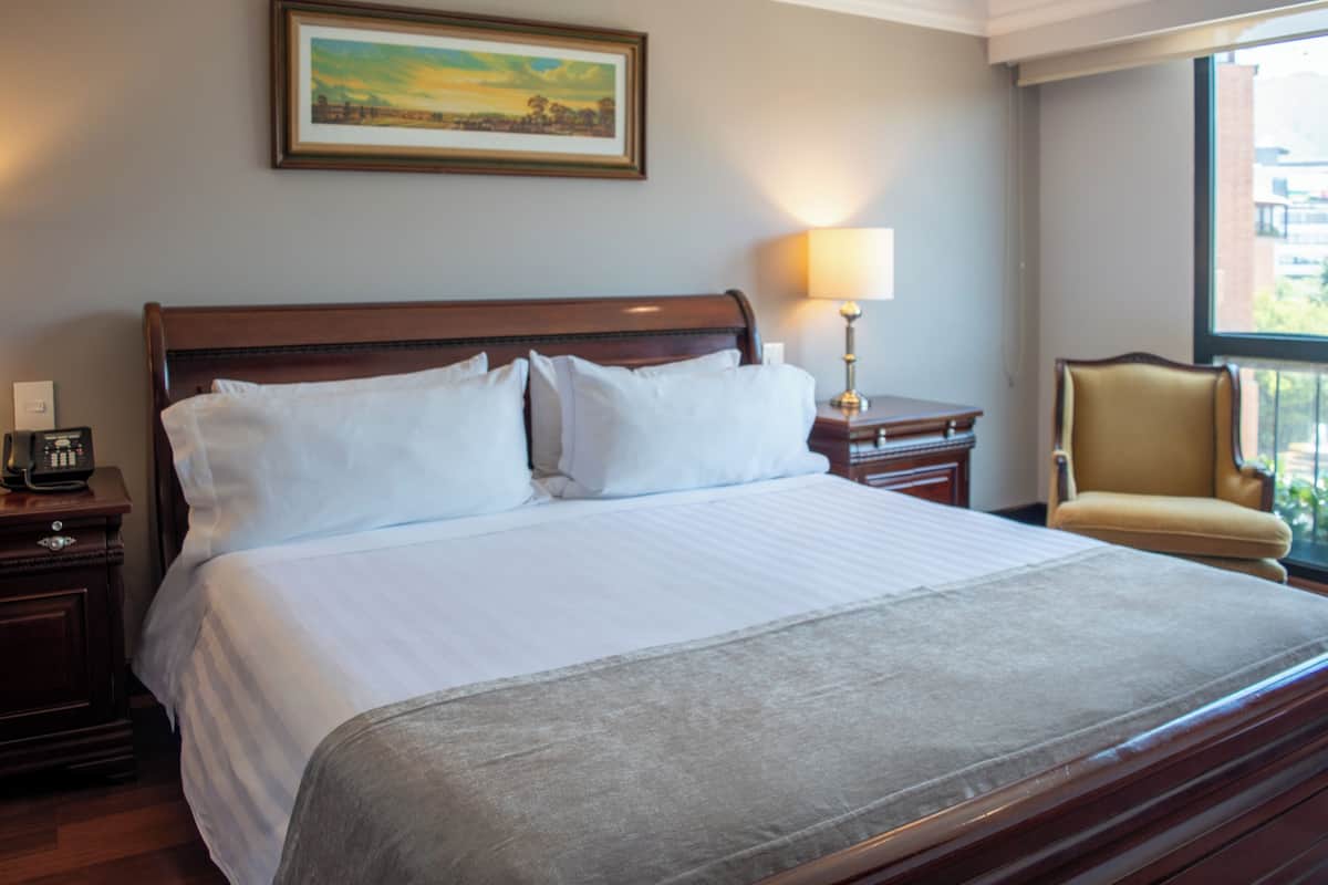 Quarto de hotel com uma cama grande, uma mesa de cabeceira com abajur, uma cadeira e uma janela com vista.