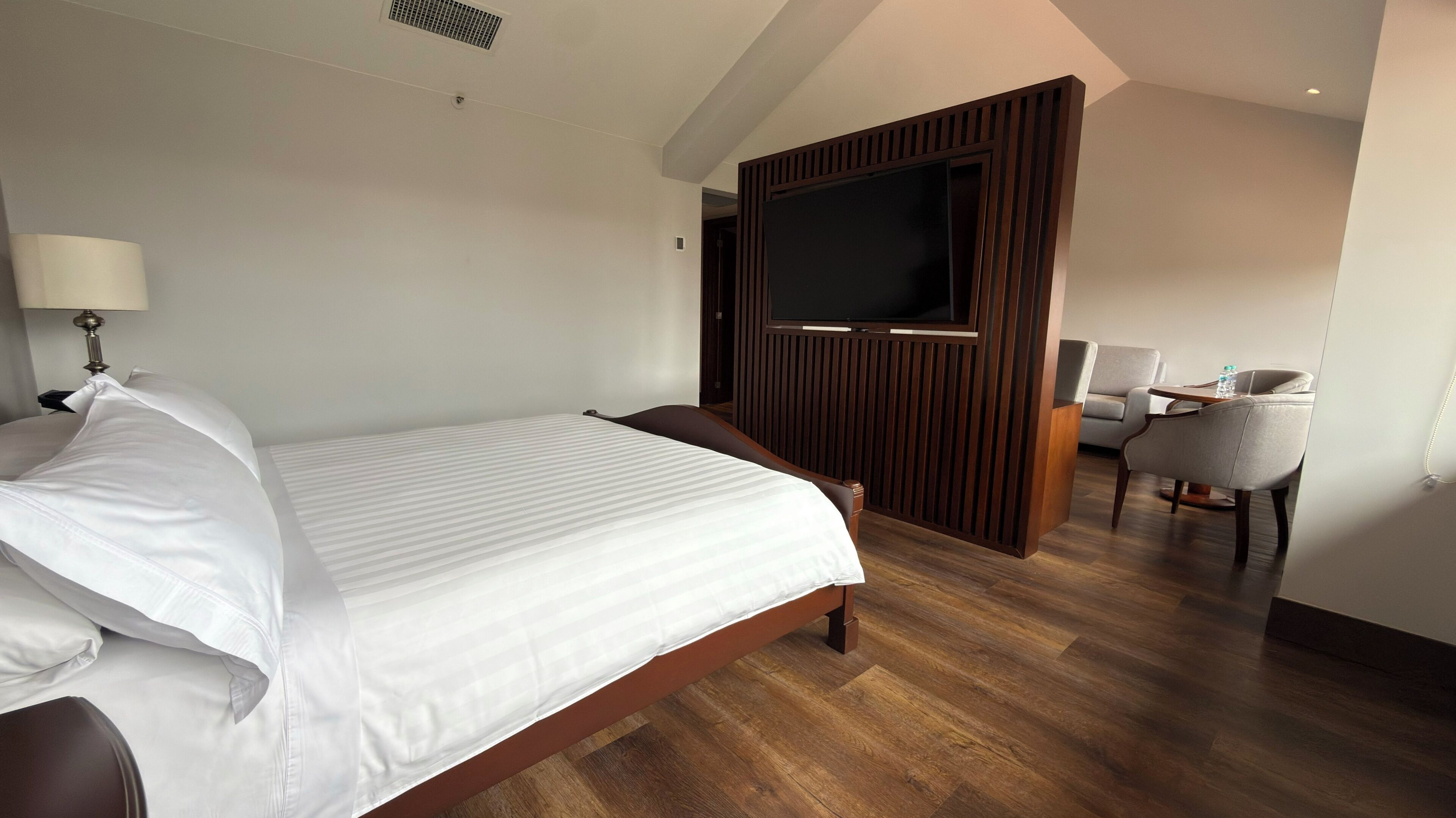 Quarto de hotel com uma cama grande, uma TV fixada em um painel de madeira, uma escrivaninha com uma cadeira e uma luminária.