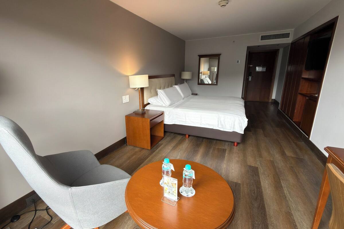 Quarto de hotel com cama, mesa de cabeceira, cadeira e uma pequena mesa redonda com garrafas de água.
