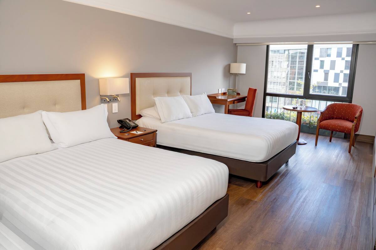 Quarto de hotel com duas camas, uma escrivaninha, uma cadeira e uma pequena mesa.