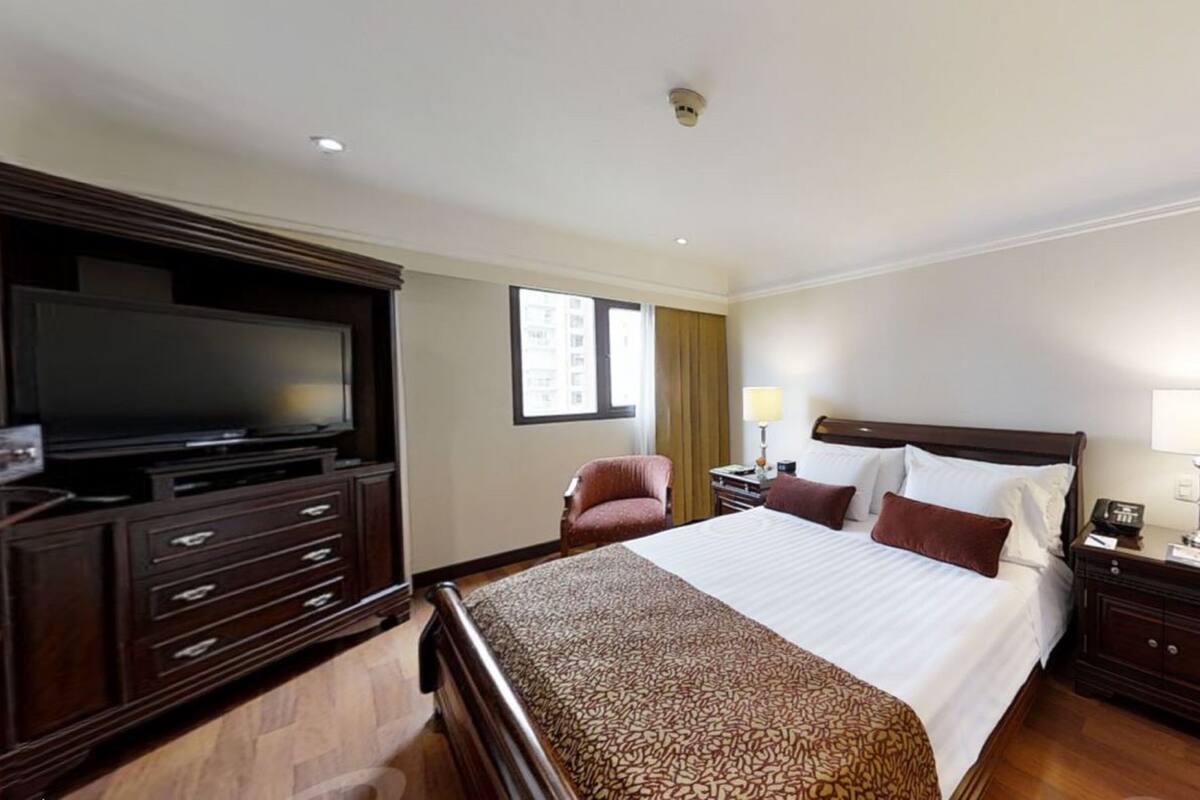 Quarto de hotel com uma cama grande, televisão, escrivaninha com abajur e uma cadeira.