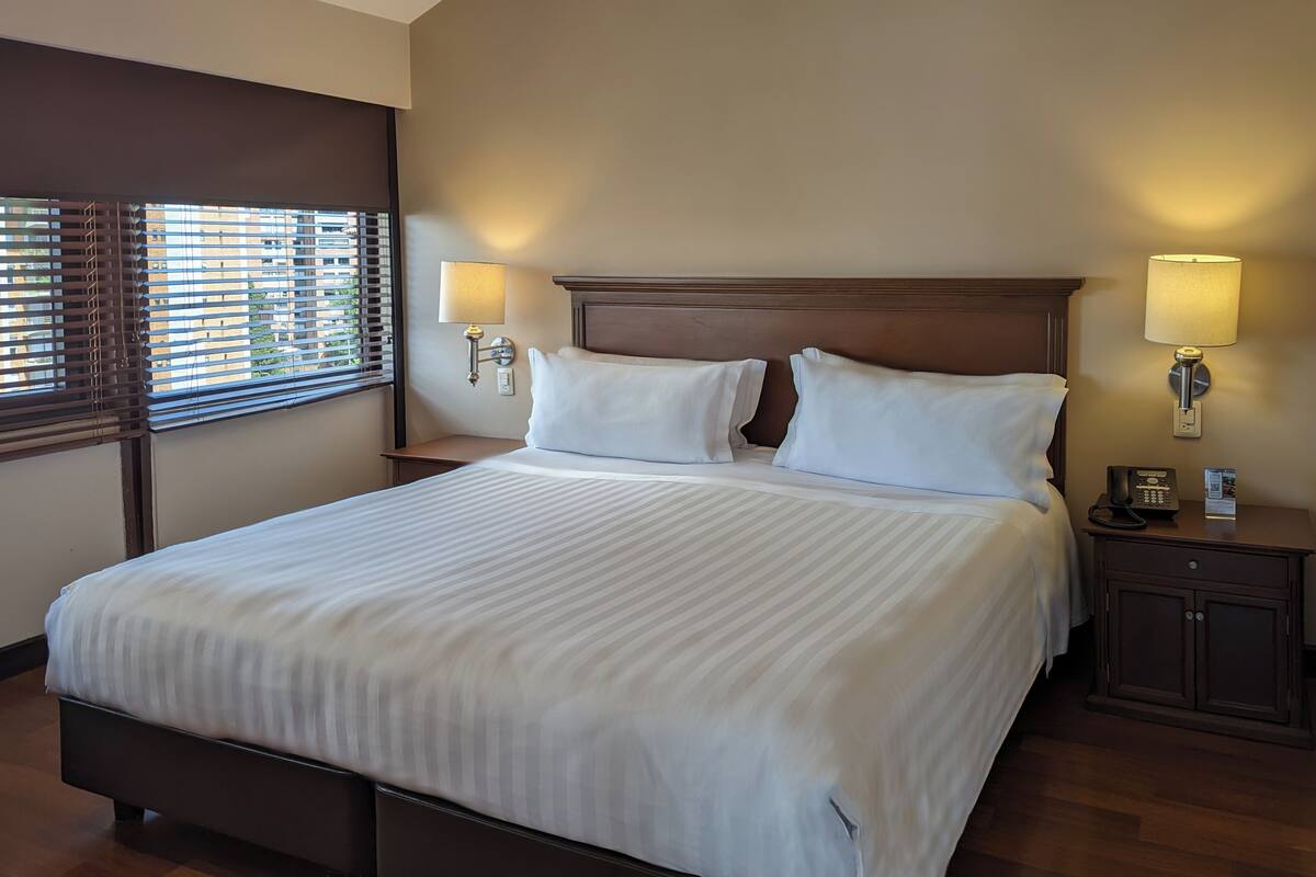 Quarto de hotel com uma cama grande, duas luminárias de cabeceira, uma mesa de cabeceira com telefone e uma janela com persianas.