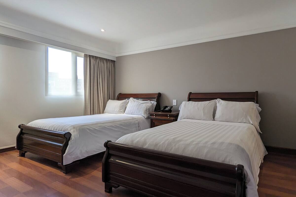 Quarto de hotel com duas camas, cabeceiras de madeira, uma mesa de cabeceira, um telefone e uma janela com cortinas.