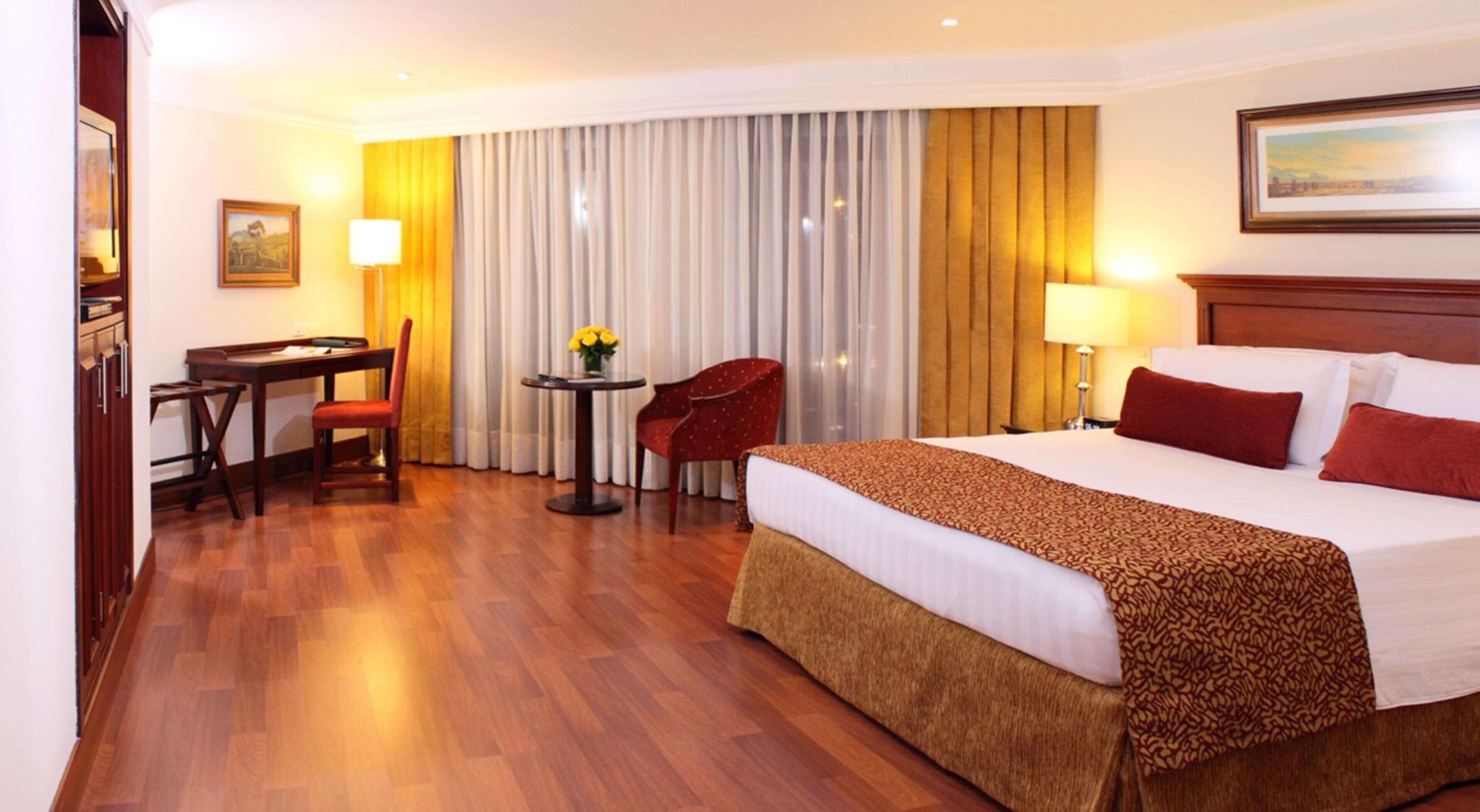 Quarto de hotel com cama, escrivaninha, cadeira, mesinha com flores e abajur.