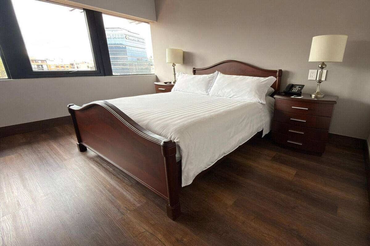 Quarto de hotel com cama de madeira, duas mesas de cabeceira com abajures, um criado-mudo e uma janela com vista para edifícios.