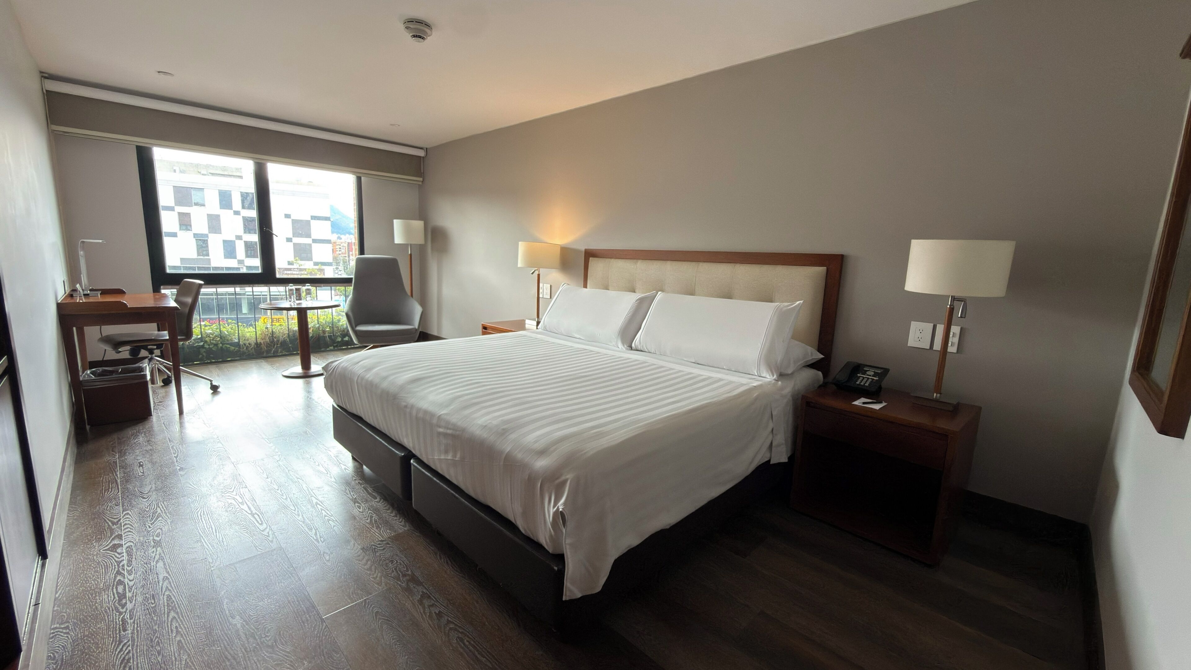 Quarto de hotel com uma cama grande, duas mesas de cabeceira com abajures, uma escrivaninha com cadeira e vista para edifícios pela janela.