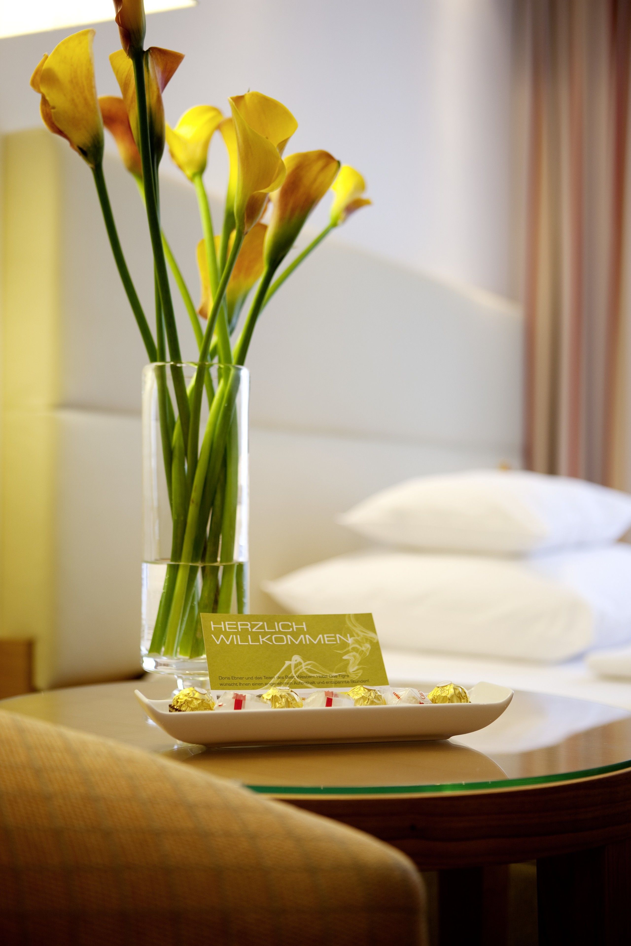 Quarto de hotel com uma cama, uma mesa de cabeceira com um vaso de flores e um prato com bombons.