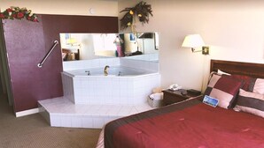 Quarto de hotel com uma cama grande, banheiro privativo com banheira, mesa de cabeceira com abajur e criado-mudo.