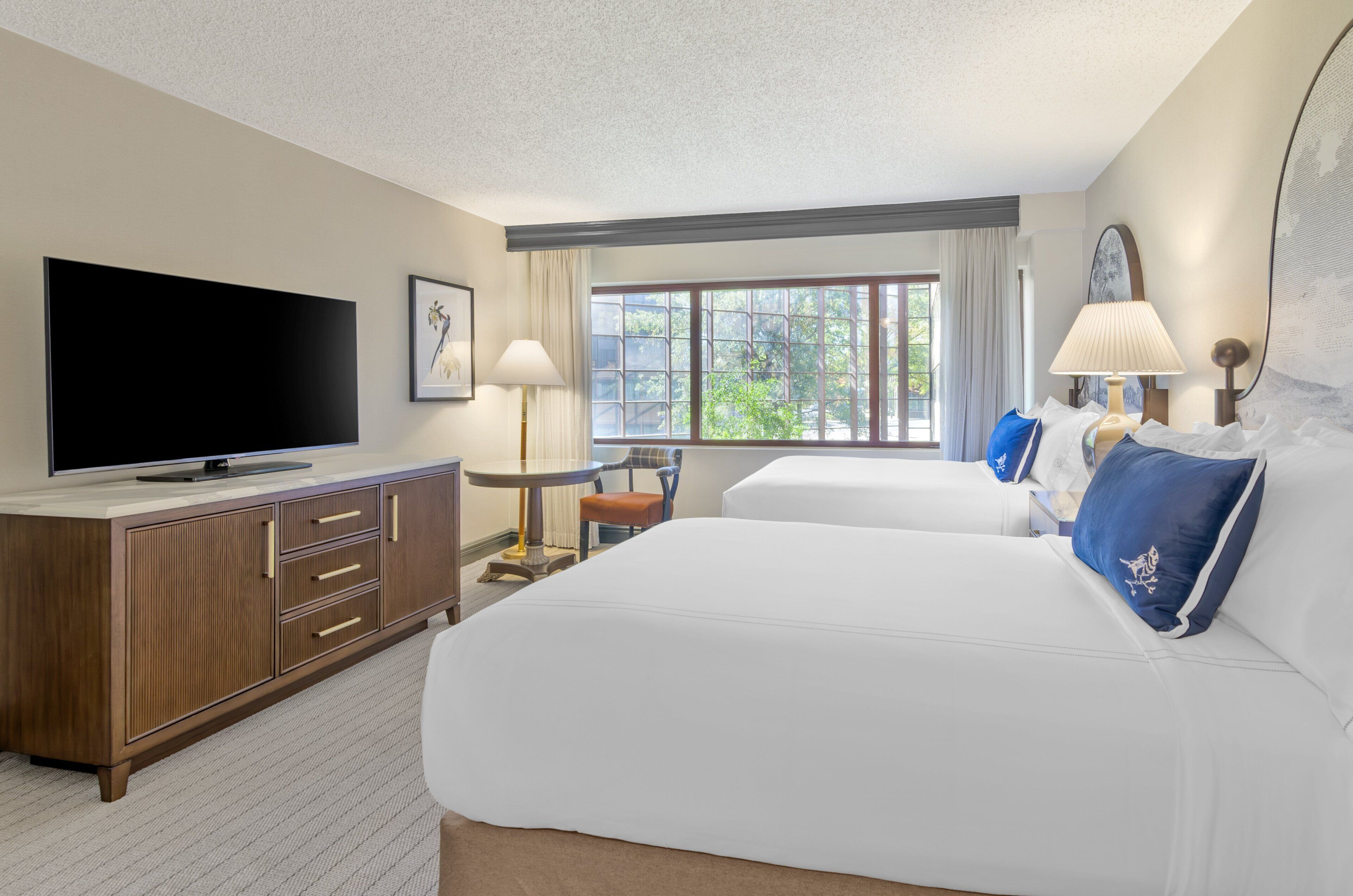 Quarto de hotel com uma cama grande, televisão, escrivaninha e janela com vista para o verde.