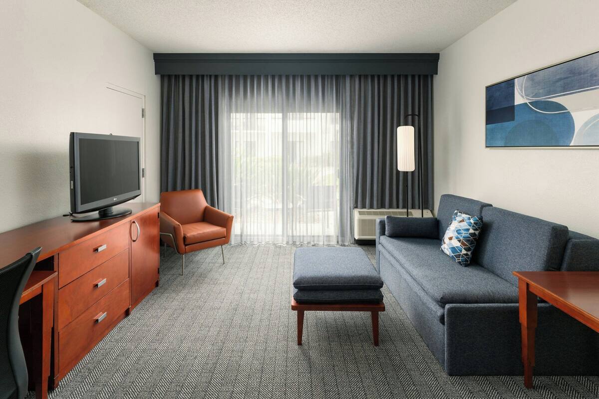 Quarto de hotel moderno com televisão de tela plana, sofá, poltrona, escrivaninha de madeira e uma janela grande com cortinas.