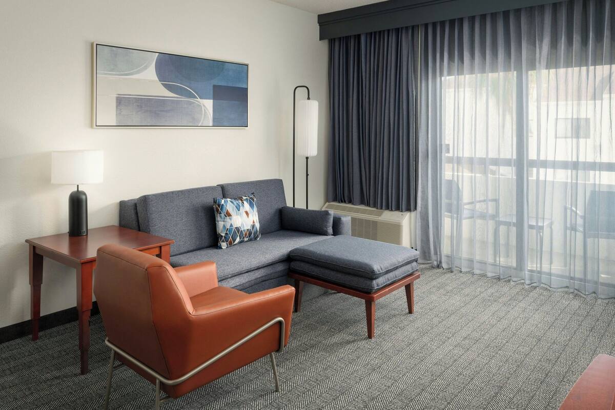 Quarto de hotel moderno com sofá cinzento, poltrona de couro marrom, mesinha e luminária de chão.