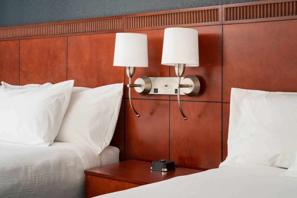 Quarto de hotel com duas camas, cabeceira de madeira, duas luminárias fixadas na parede e um relógio digital na mesinha de cabeceira.