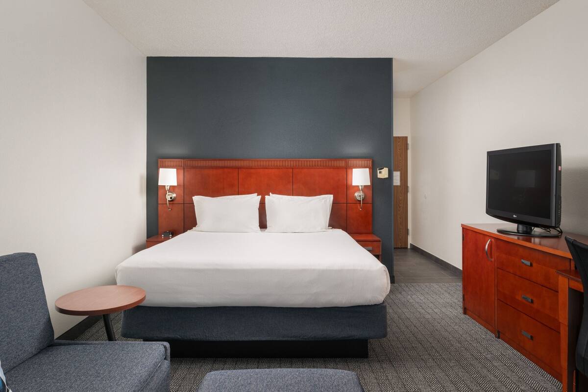 Quarto de hotel com uma cama grande, duas mesas de cabeceira com luminárias, uma televisão sobre um móvel de madeira e uma pequena mesa redonda.