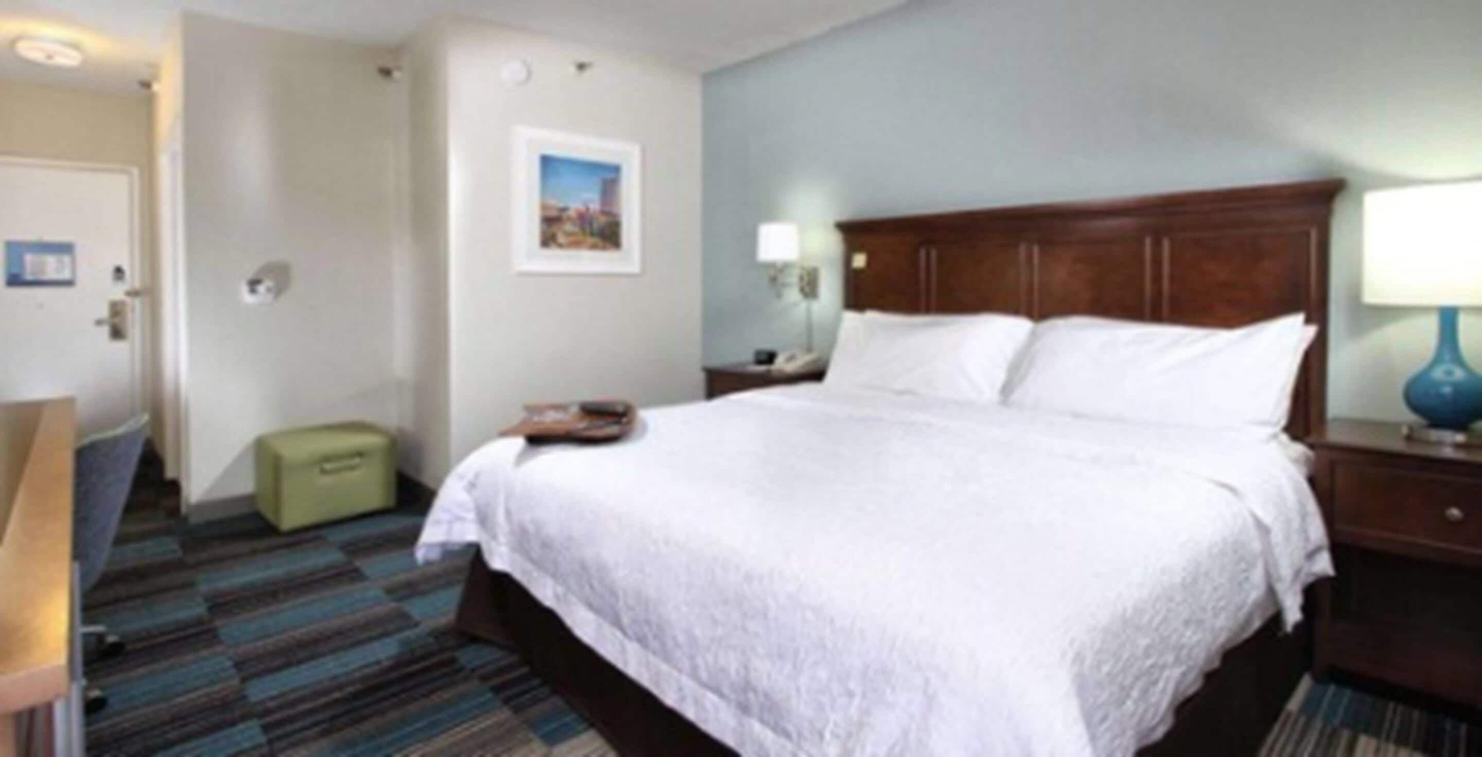 Quarto de hotel com uma cama grande, uma mesinha de cabeceira com luminária, uma pequena mesinha com telefone, um quadro na parede e uma mala no chão.