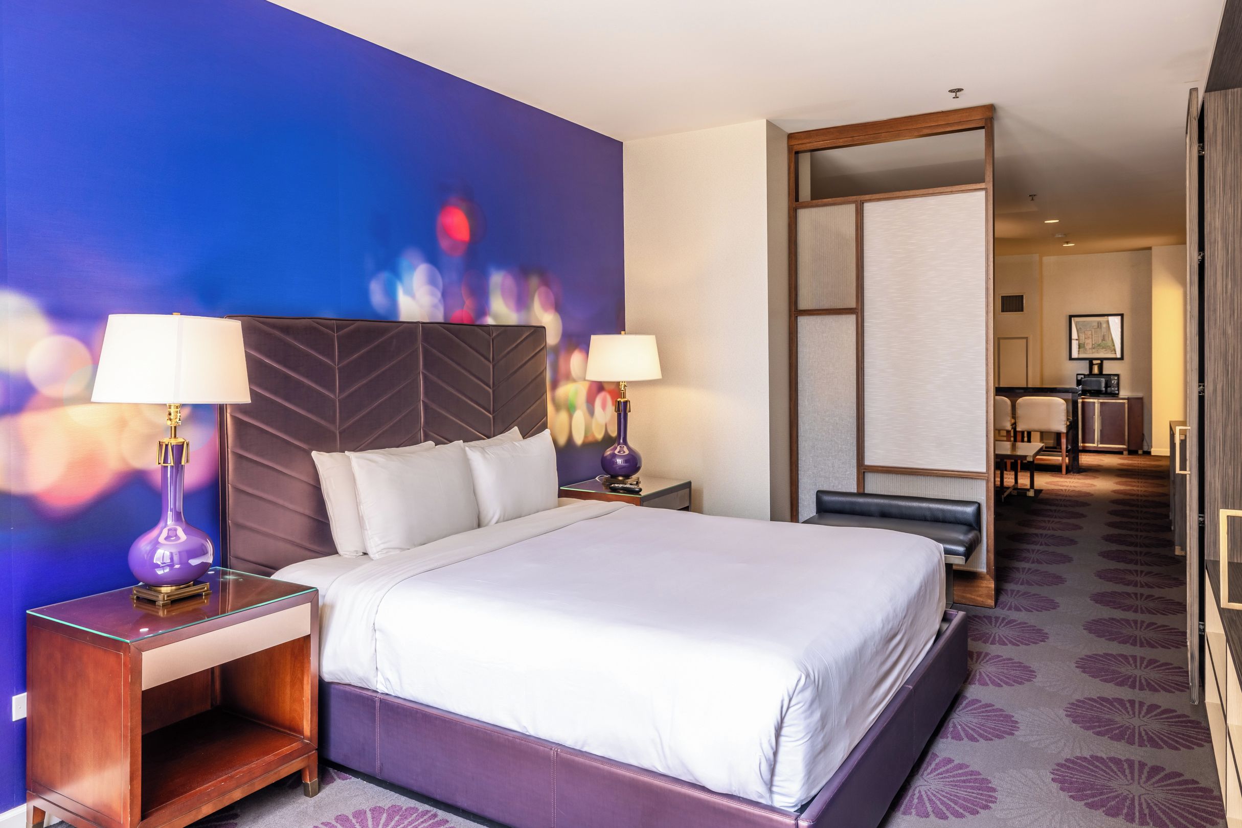 Quarto de hotel com uma cama grande, duas mesas de cabeceira com luminárias, uma cabeceira e vista para o corredor.