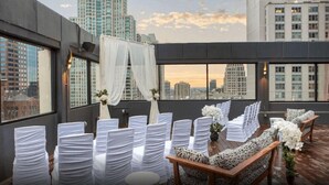 Espaço no terraço com cadeiras brancas e um sofá, adornado com tecido branco e arranjos florais, com vista para a paisagem urbana.
