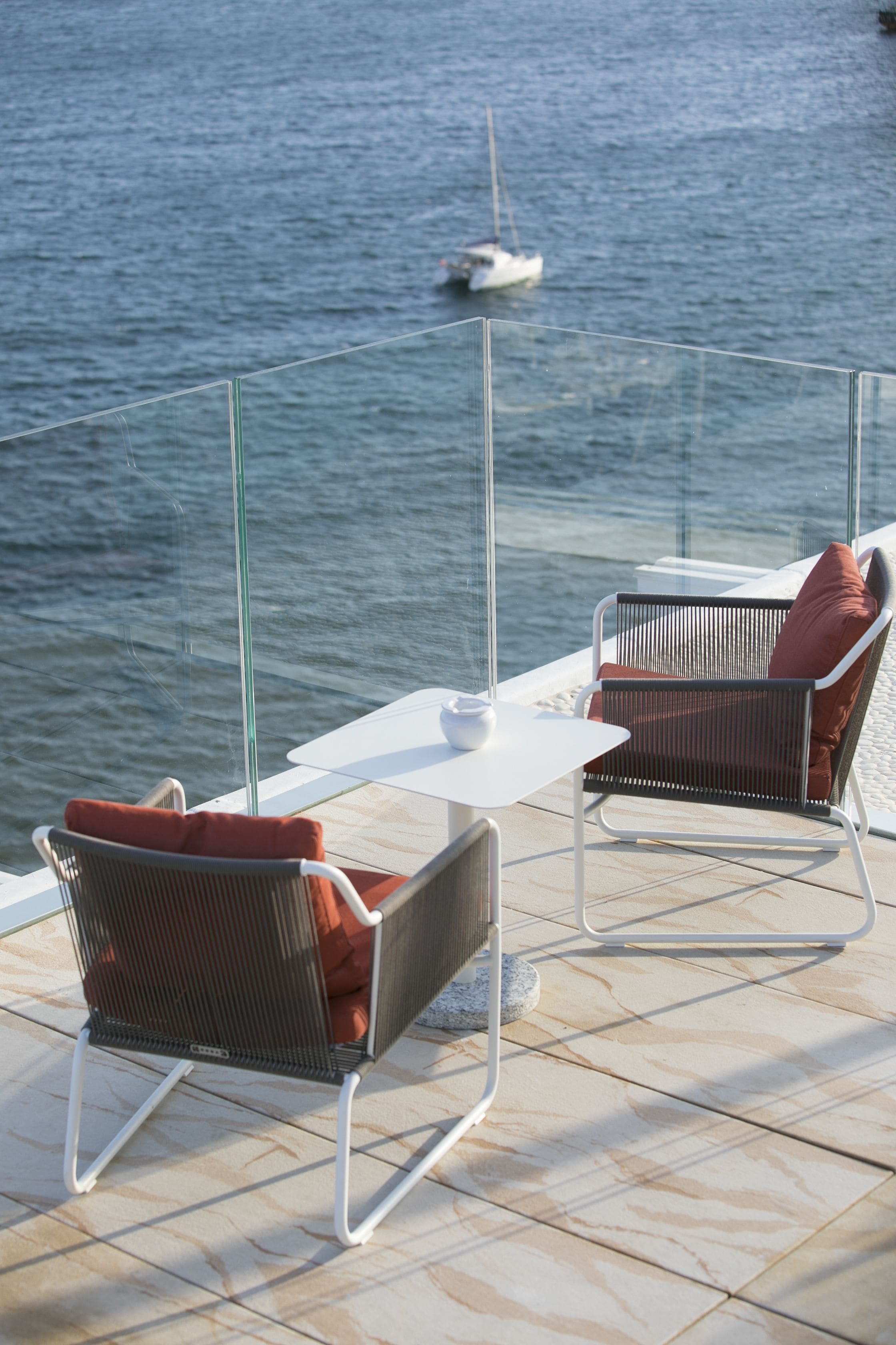 Terraço com duas cadeiras e uma mesa, com vista para um corpo d'água onde se vê um veleiro ao longe.