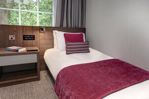 Quarto de hotel com uma cama, uma mesa de cabeceira com um livro, um controle remoto e uma janela com cortinas.