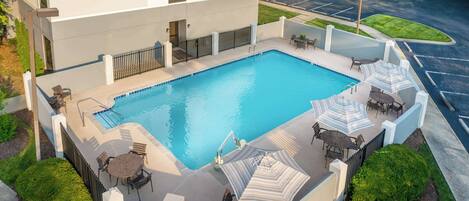 Uma área de piscina no terraço com uma piscina retangular, rodeada por espreguiçadeiras e guarda-sóis, e vista para um estacionamento e áreas verdes.