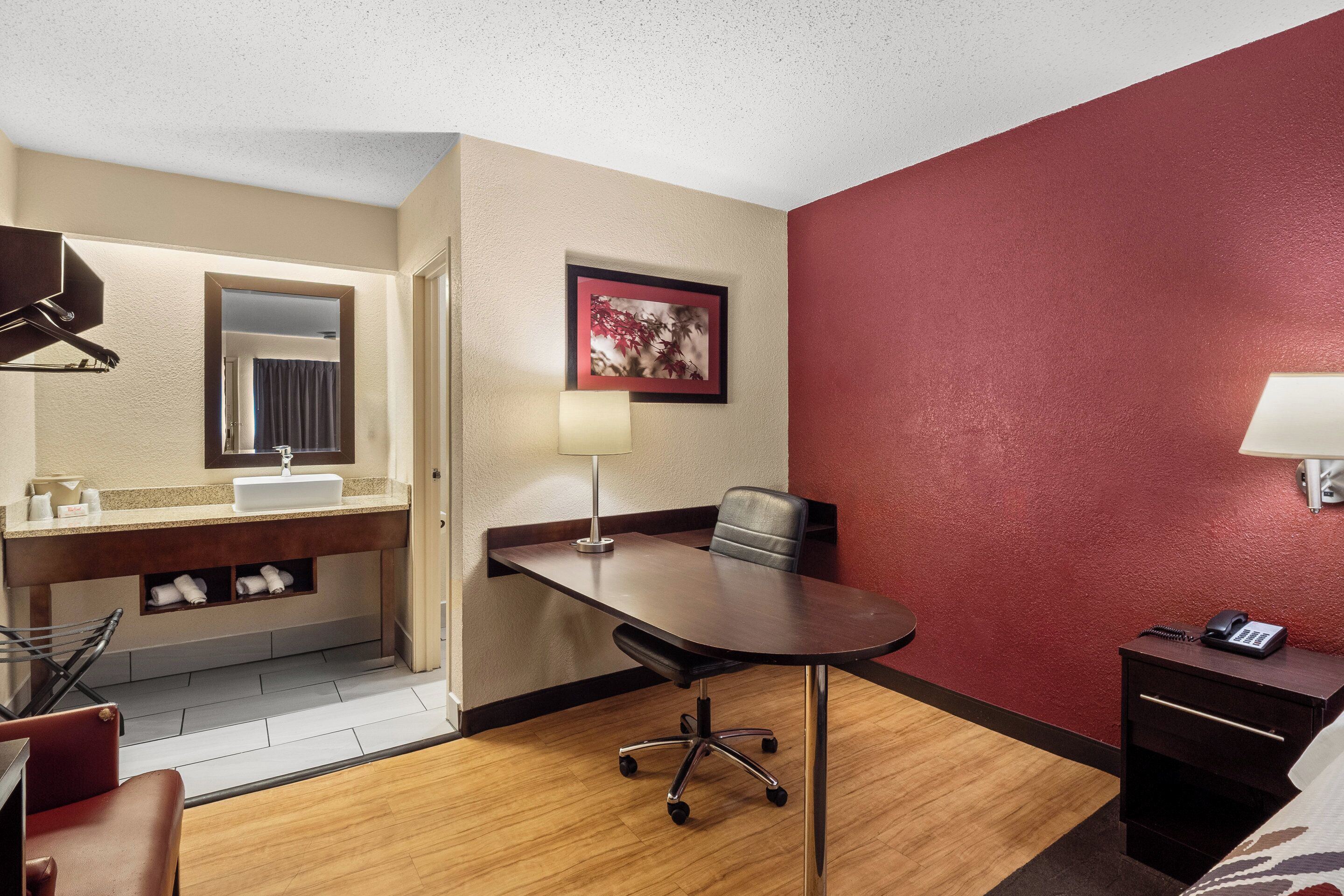 Quarto de hotel com piso de madeira, uma escrivaninha com cadeira, uma pequena mesa de cabeceira com abajur, telefone e uma cama.
