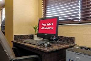 Mesa com monitor de computador exibindo 'Wi-Fi Gratuito em Todos os Quartos', teclado, mouse, impressora e planta em vaso.