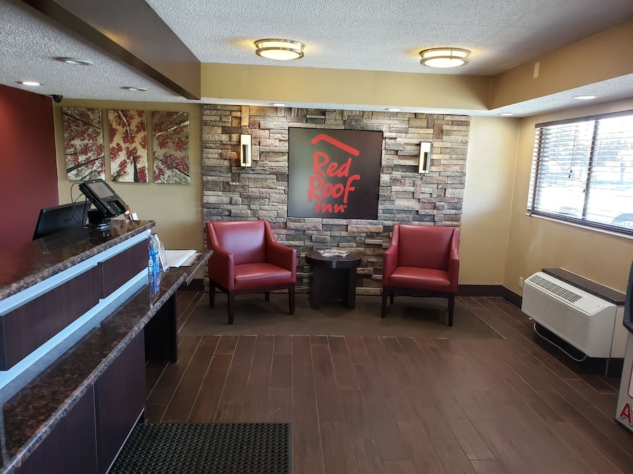 Área de recepção do Red Roof Inn com parede de pedra ao fundo, duas poltronas de couro vermelho, balcão de recepção com terminal de pagamento e uma janela com persianas.