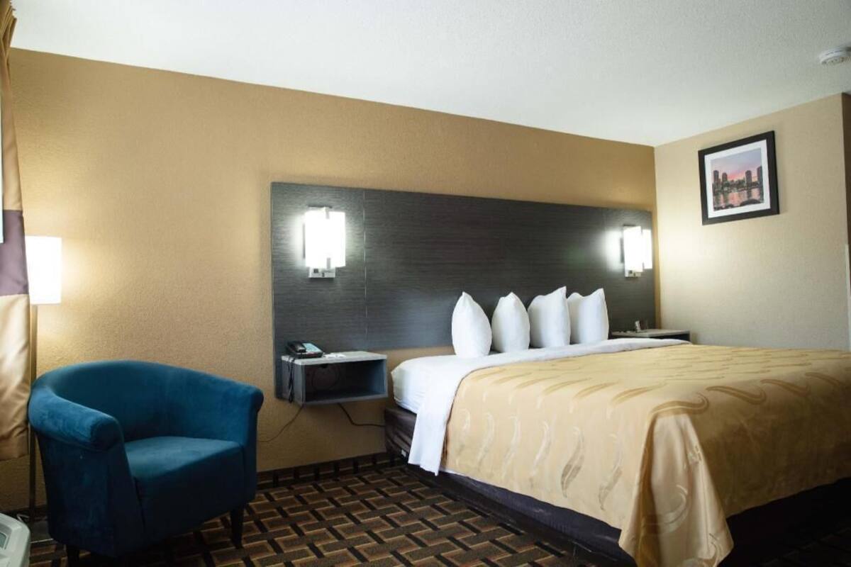 Quarto de hotel com uma cama, uma cadeira, uma mesinha de cabeceira e uma luminária fixada na parede.