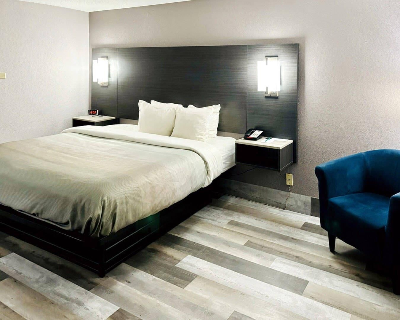 Quarto de hotel moderno com uma cama grande, mesas de cabeceira, um banquinho e um candeeiro fixado na parede.
